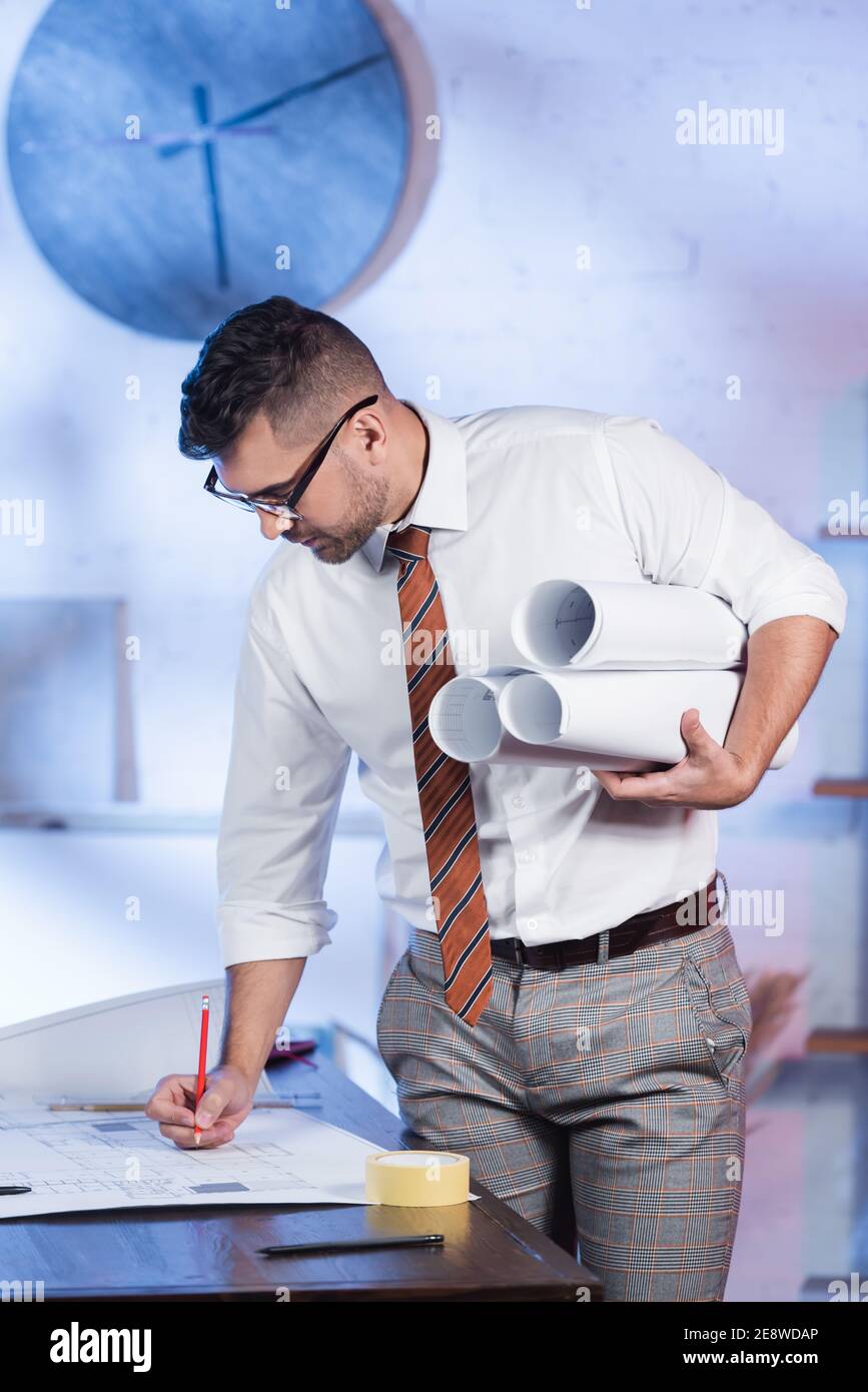 architect writing on blueprint while holding rolled plans in office ...