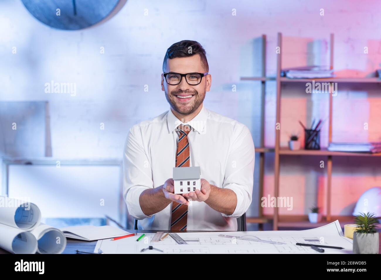 happy architect holding house model near blueprints on desk in office ...