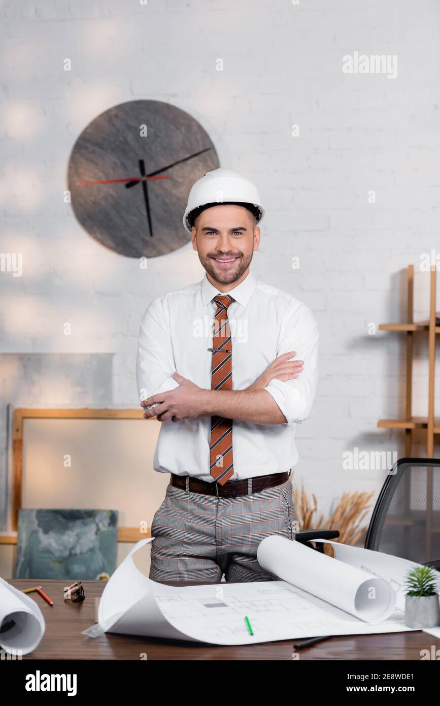 happy architect in helmet standing with crossed arms near blueprints on ...