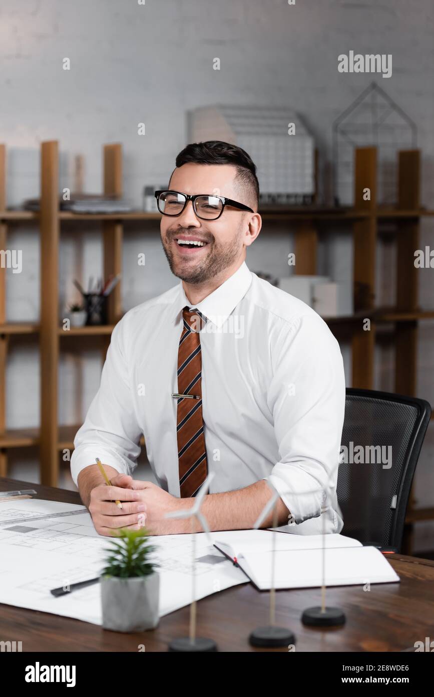 excited architect laughing at workplace near blueprint, blurred ...