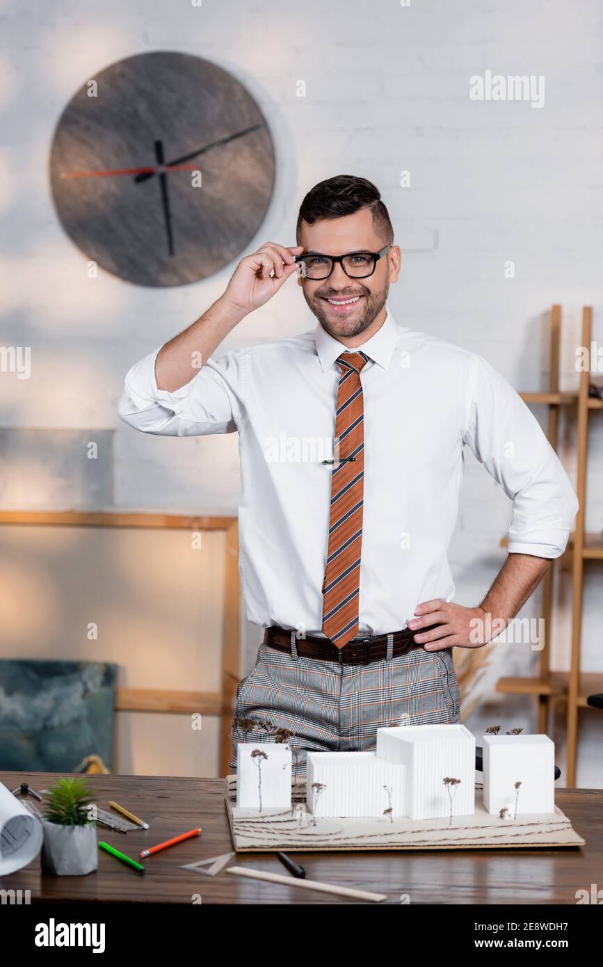 happy architect looking at camera while standing with hand on hip near ...