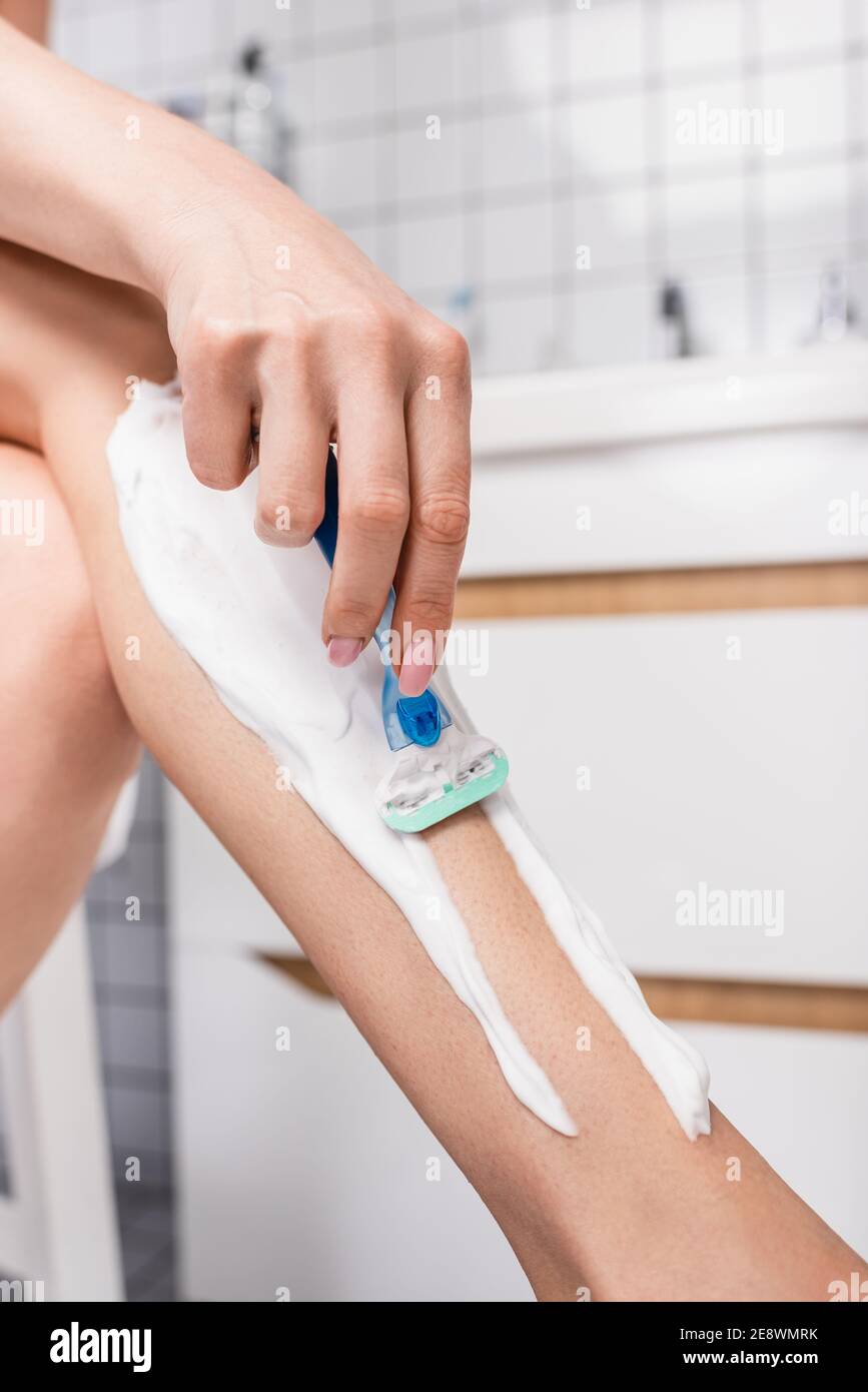 partial view of woman with shaving cream shaving leg with safety razor ...
