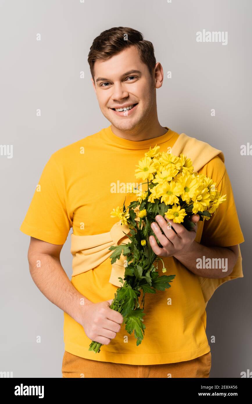 Young man smiling at camera while holding yellow chrysanthemums on grey ...