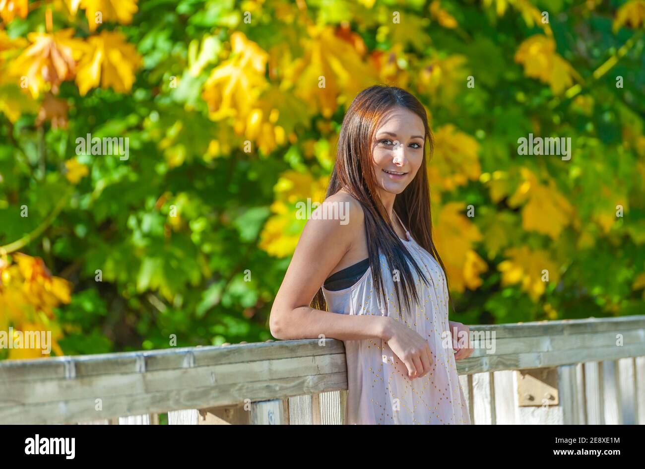 A young woman poses along the railing of a bridge with fall leaves in ...