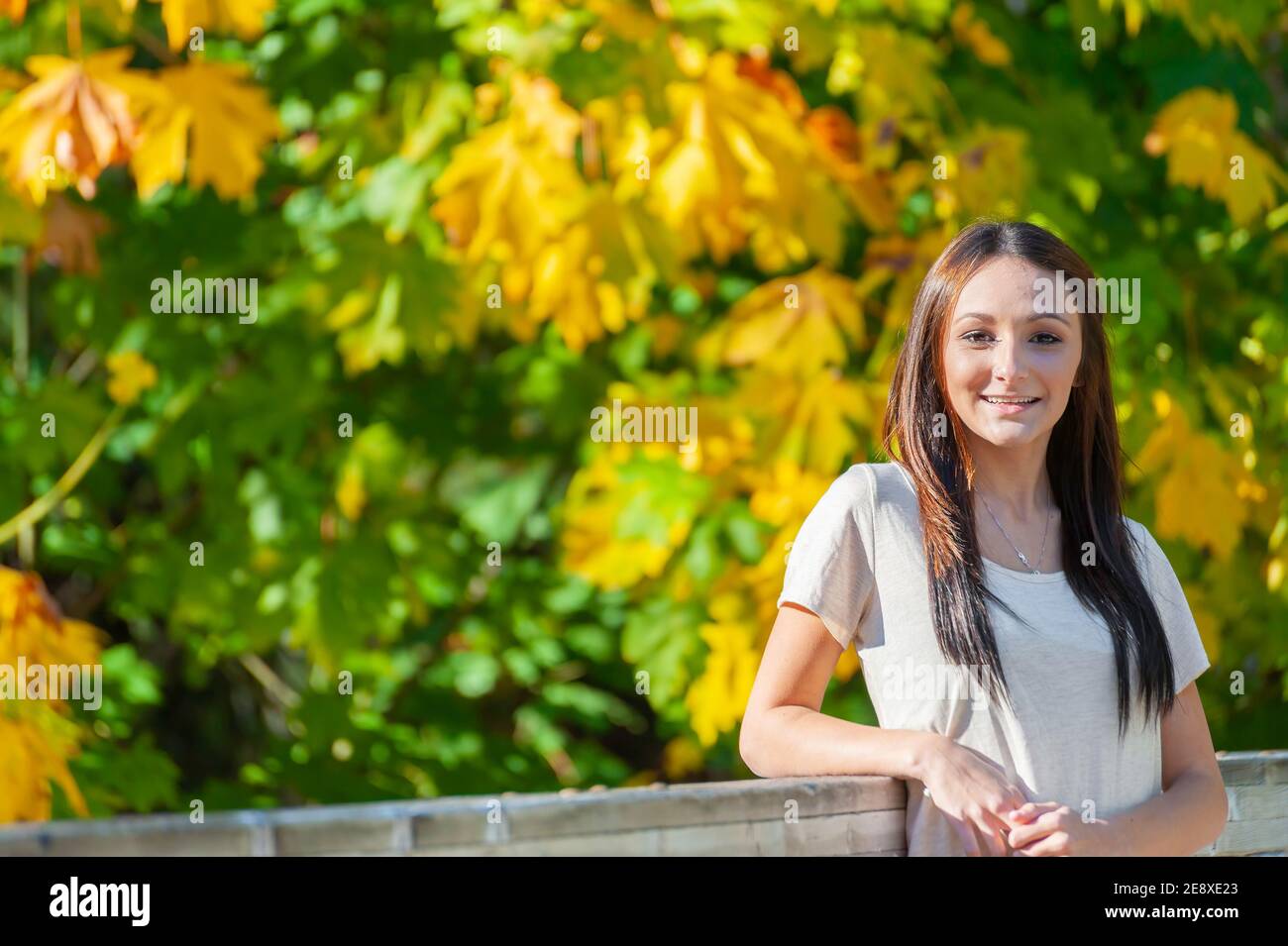 A young woman poses along the railing of a bridge with fall leaves in ...