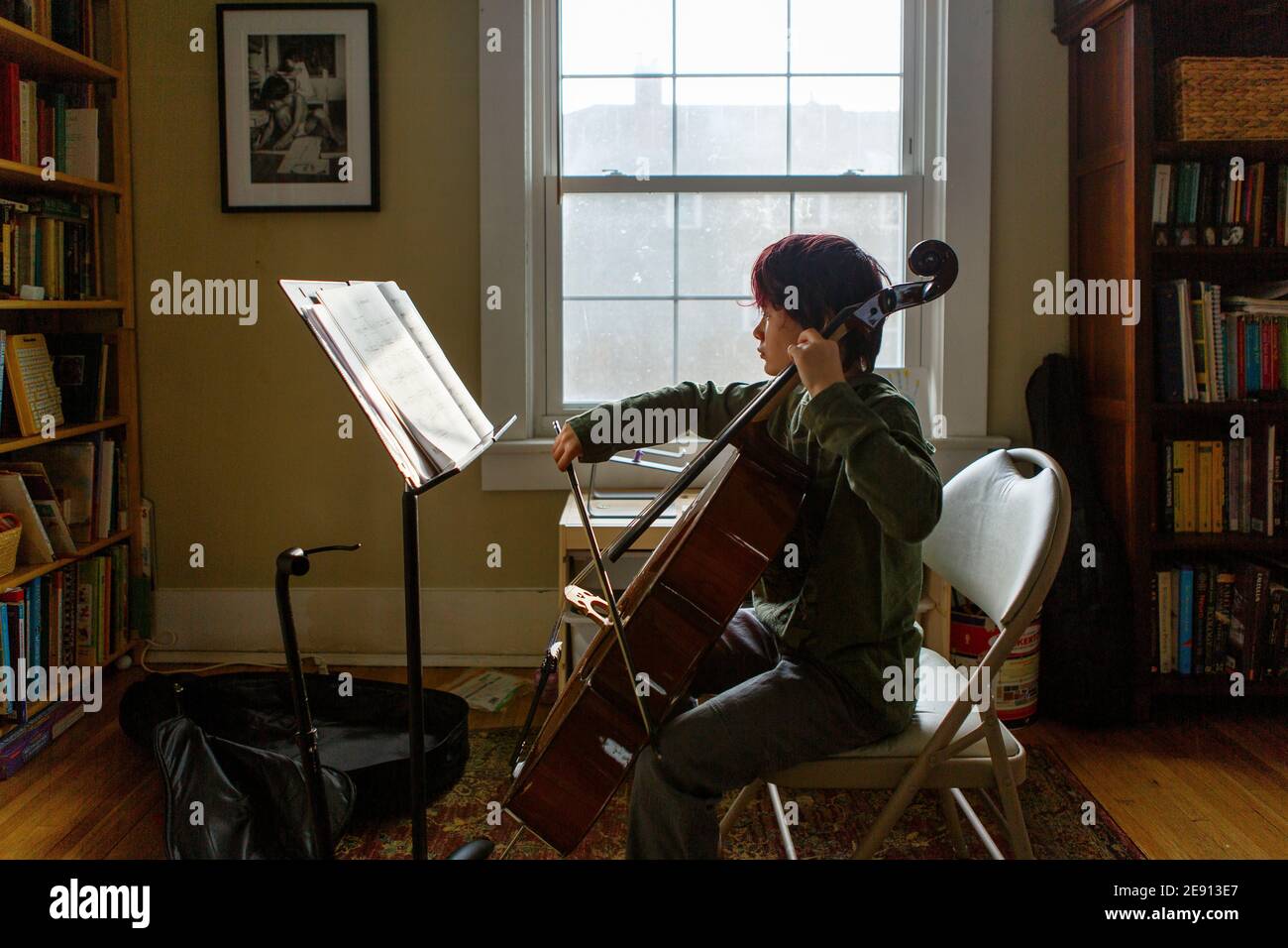 side-view of boy sitting in front of window practicing cello in sun ...