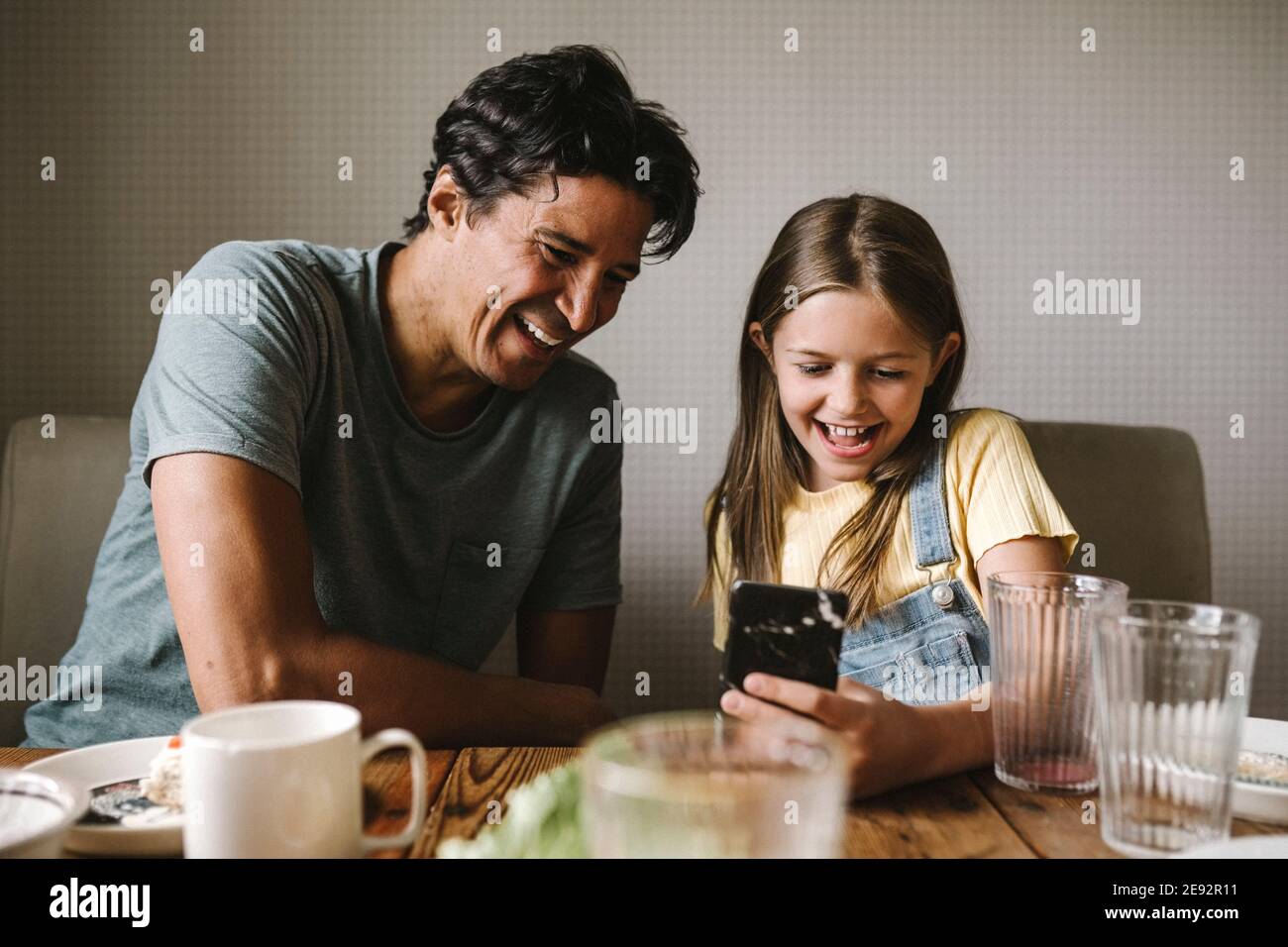 Smiling father and daughter using mobile phone at dining table Stock ...