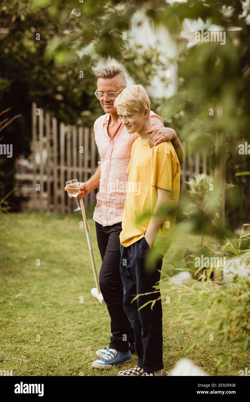 Smiling father with arm around son holding wineglass while standing in ...