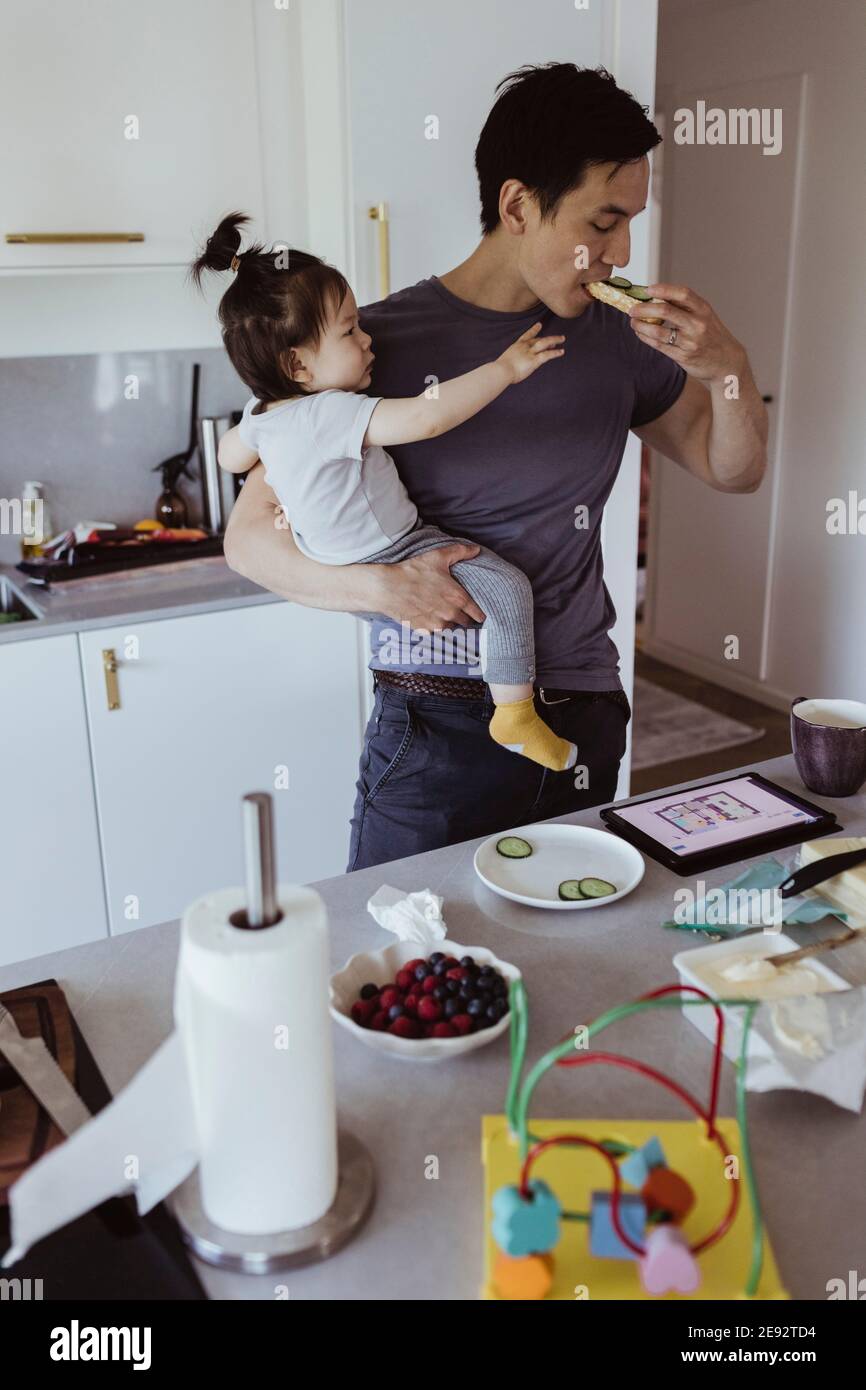 Father eating food while baby son looking at him in kitchen at home ...
