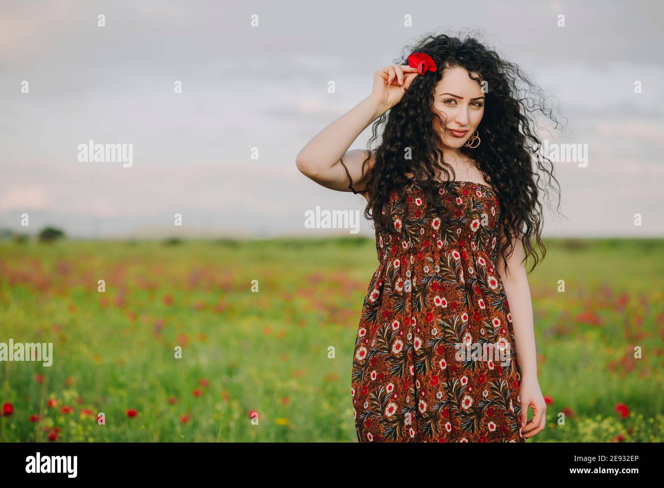 A curly hair girl wears a wild poppy flower in her hair in green field ...