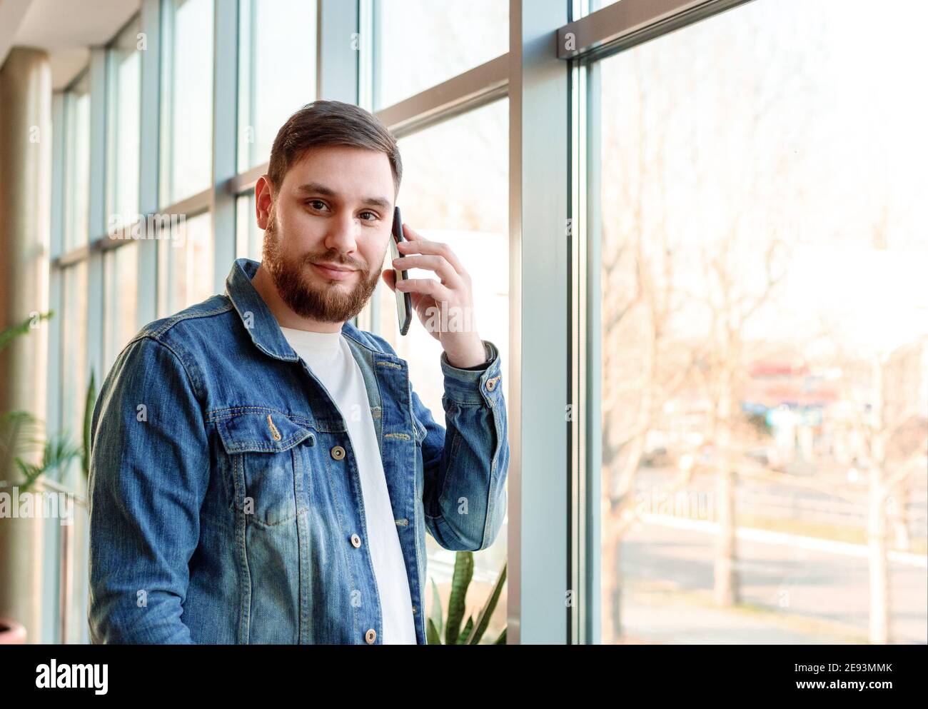 Young man making call, talking by phone indoors near window wall in ...