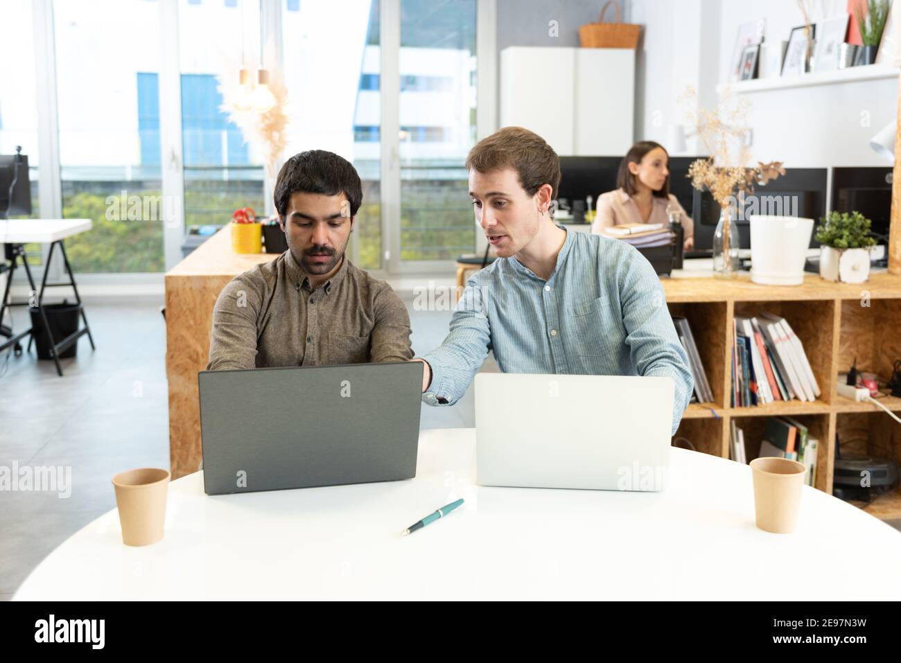 Two male coworkers working on their laptops helping each other Stock ...