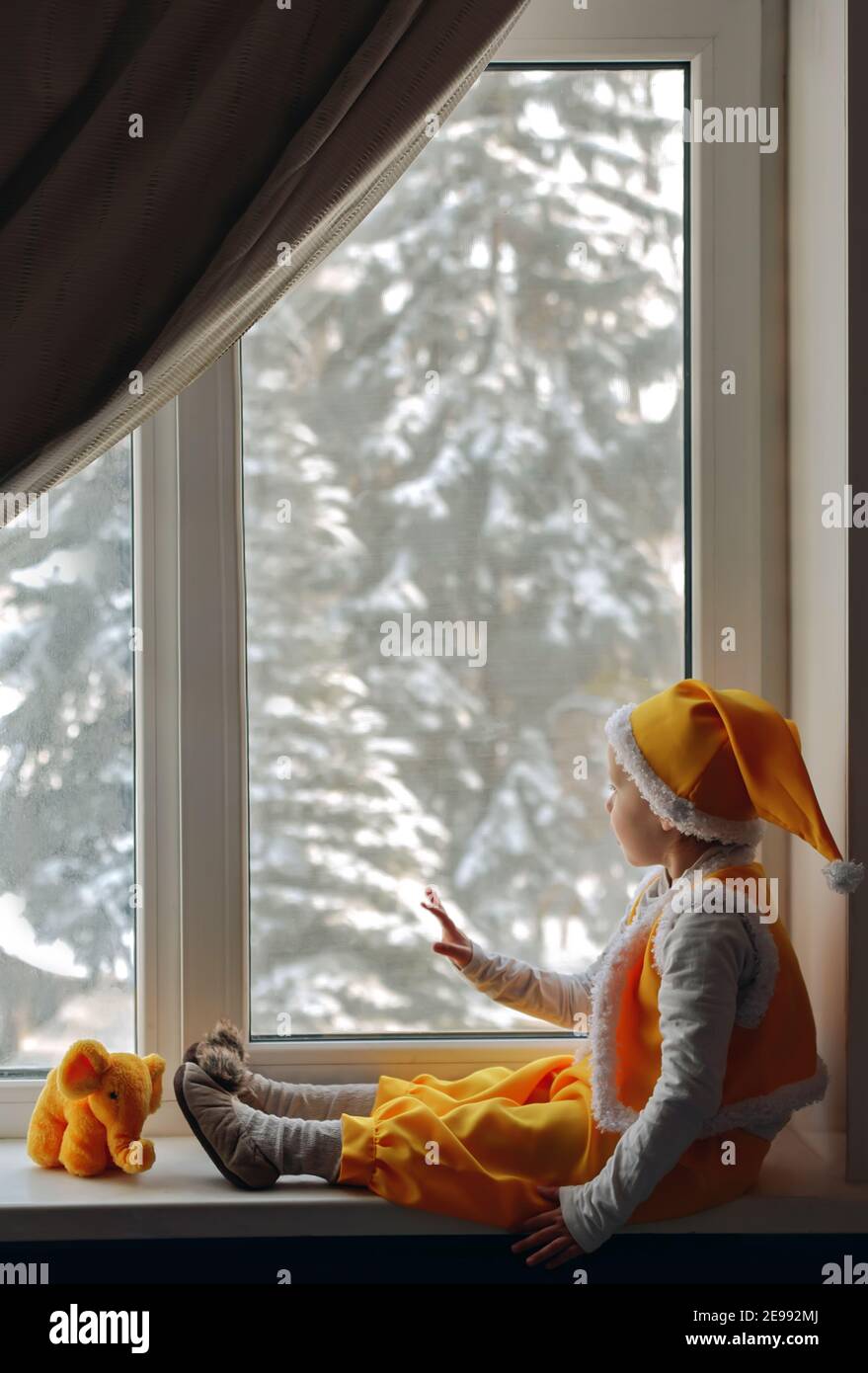 Boy sit at window and put hand on glass in cute yellow hat with toy ...