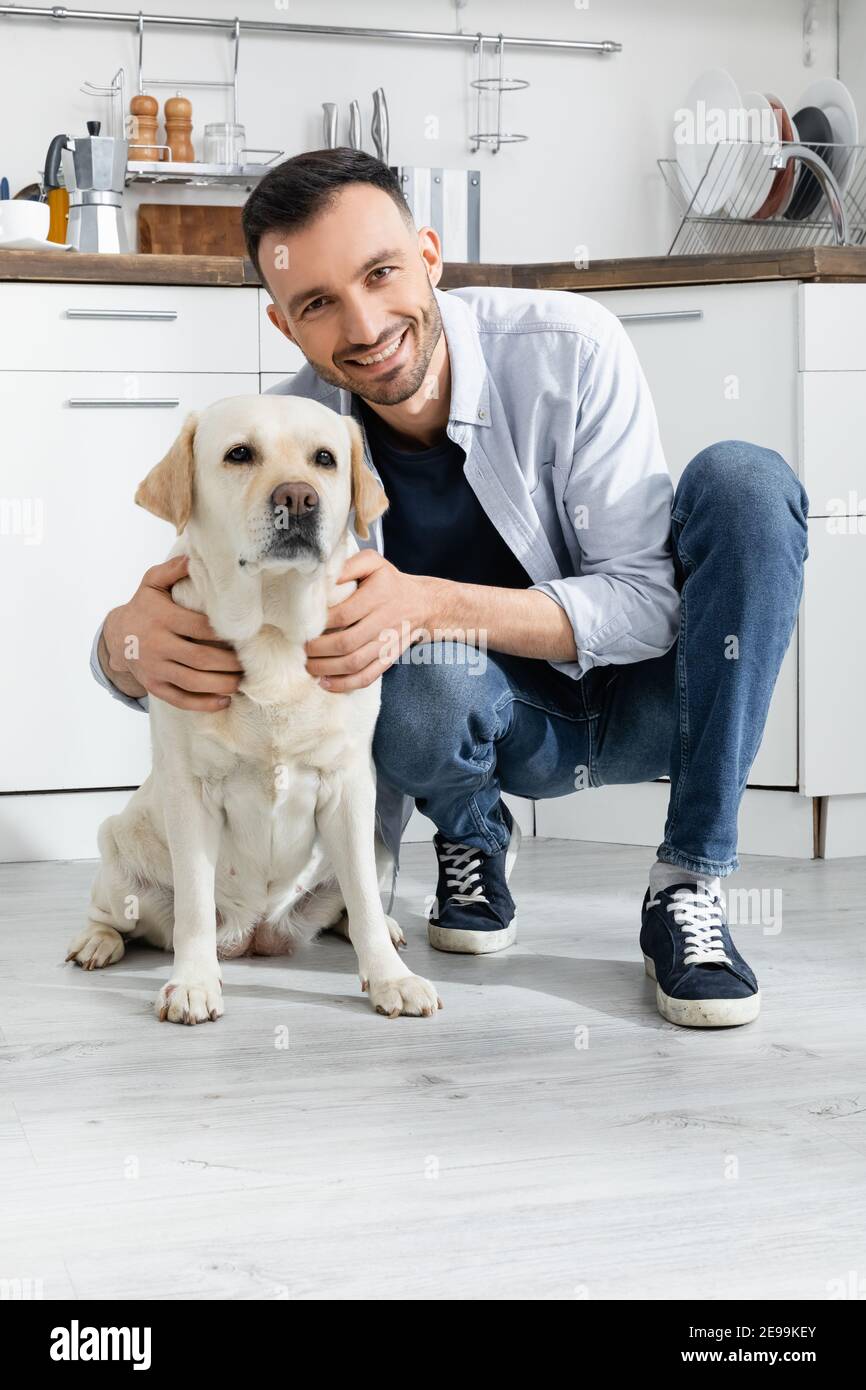 cheerful man in jeans sitting and cuddling labrador at home Stock Photo ...