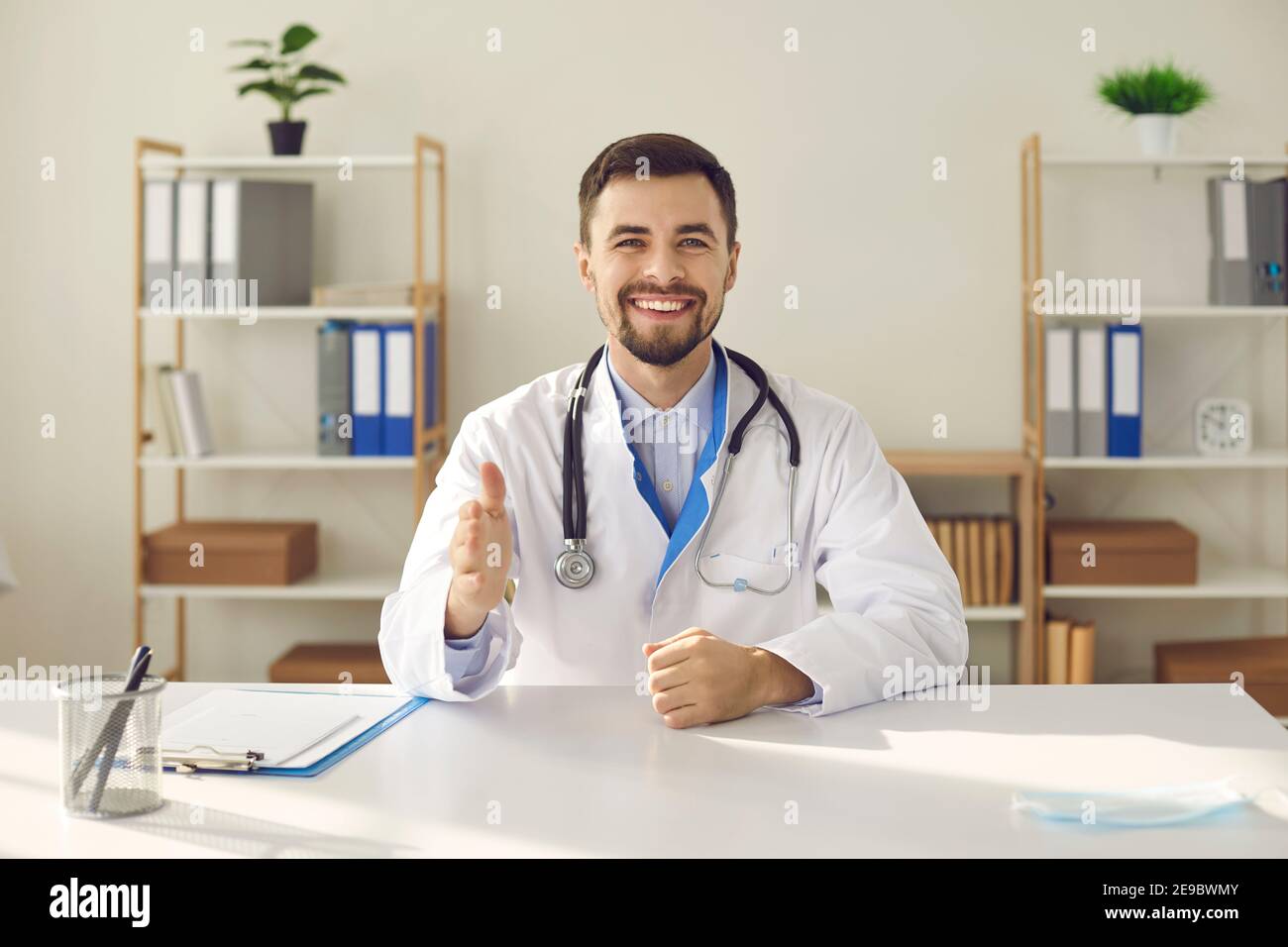 Happy young doctor sitting at his desk, smiling and reaching out for a ...