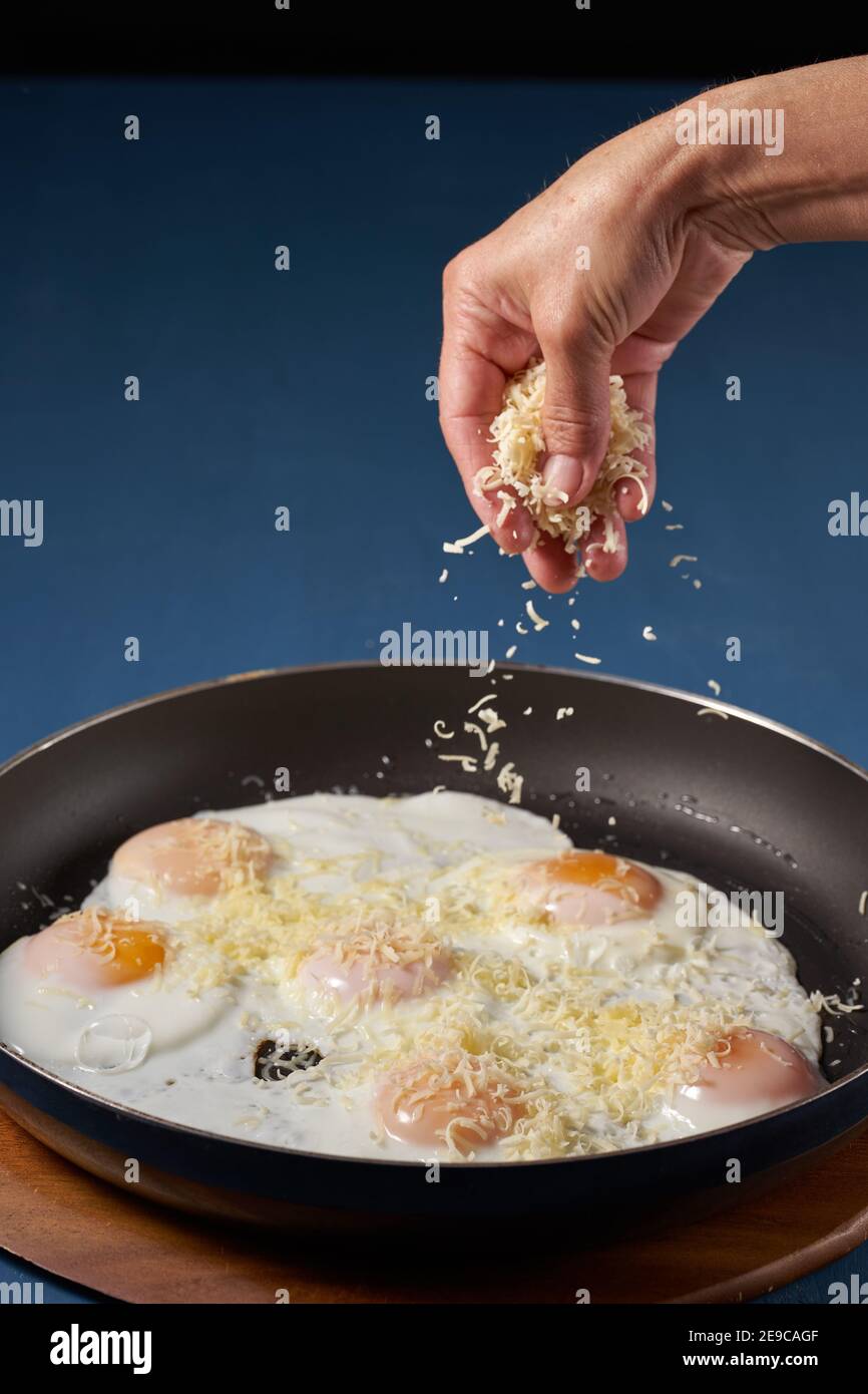 Woman sprinkling grated cheddar cheese over frying eggs in the pan ...