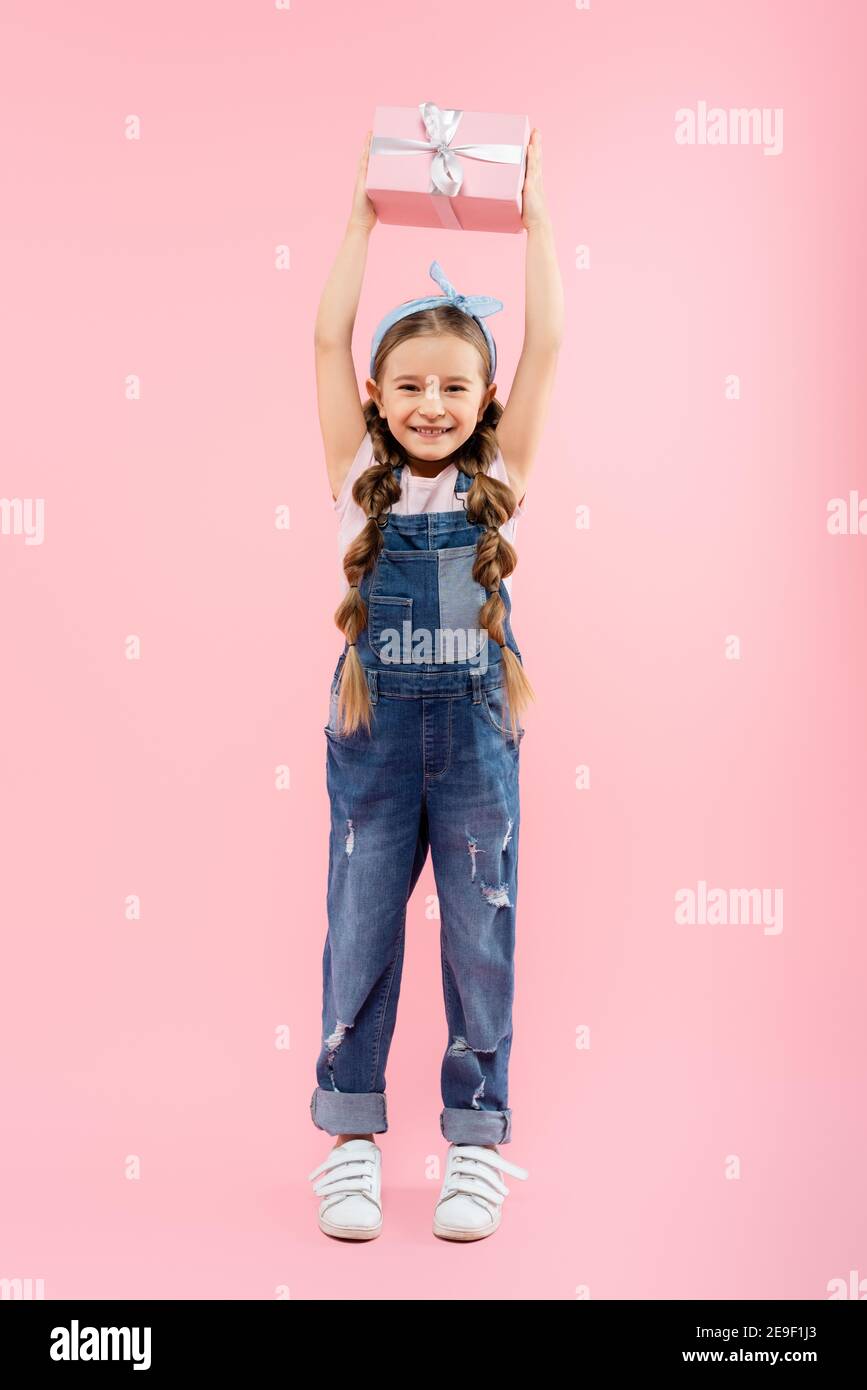 full length of happy kid smiling and holding present above head on pink ...
