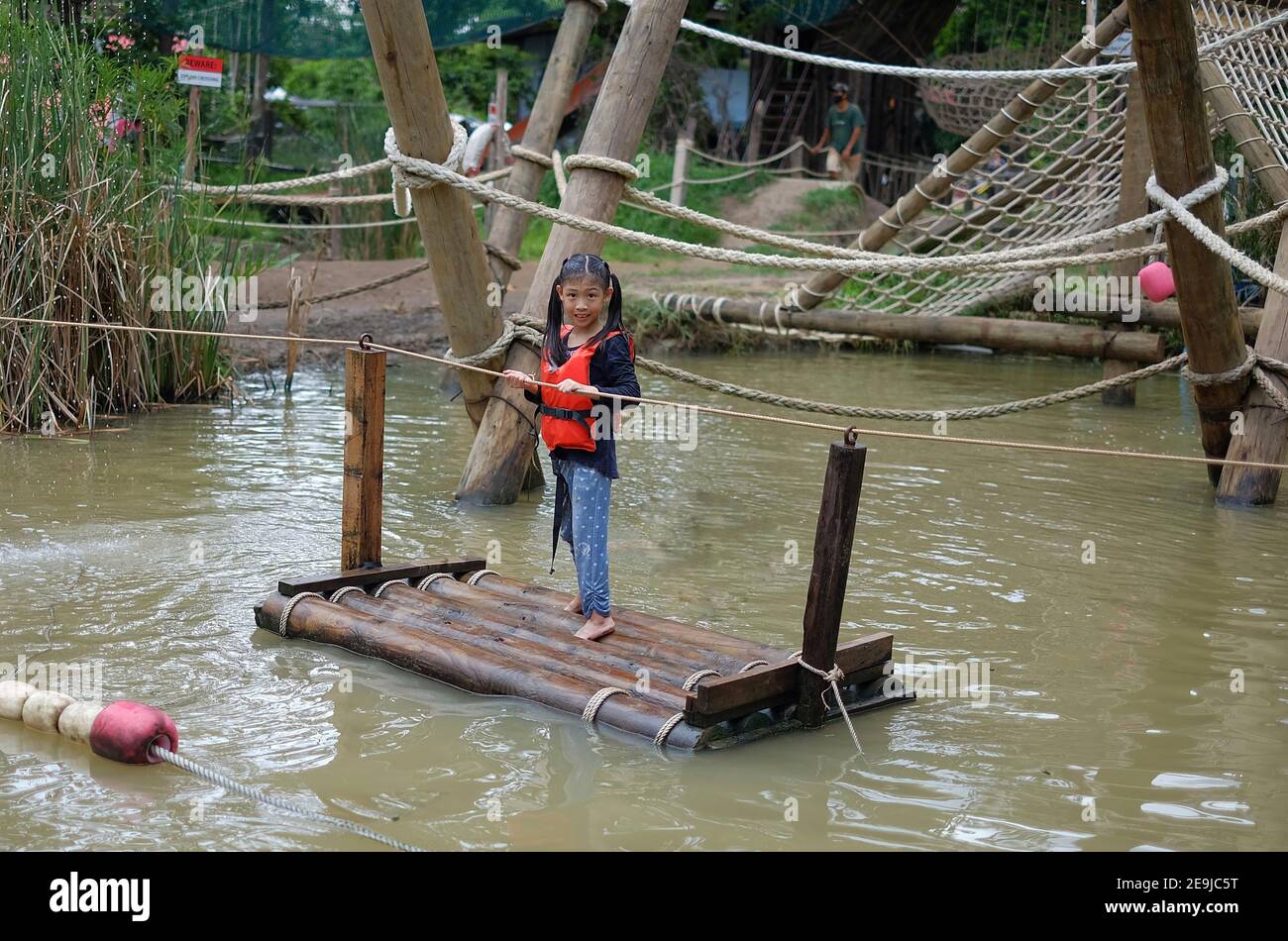 A cute young Asian girl, wearing an orange life vest, is using a bamboo ...