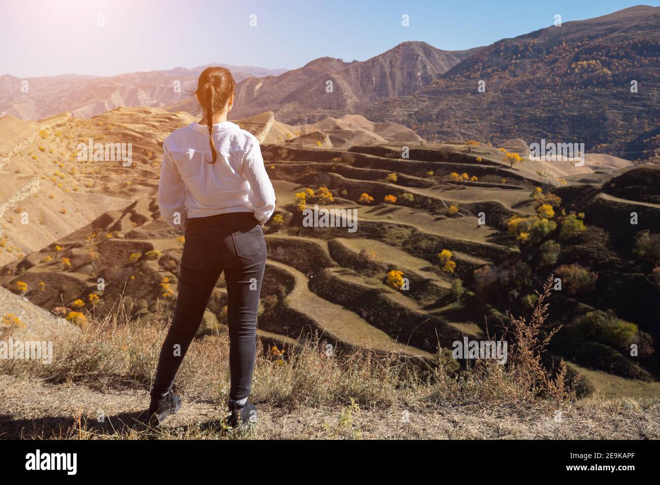 Brunette in white blouse and black denim jeans stands on brown hill top ...