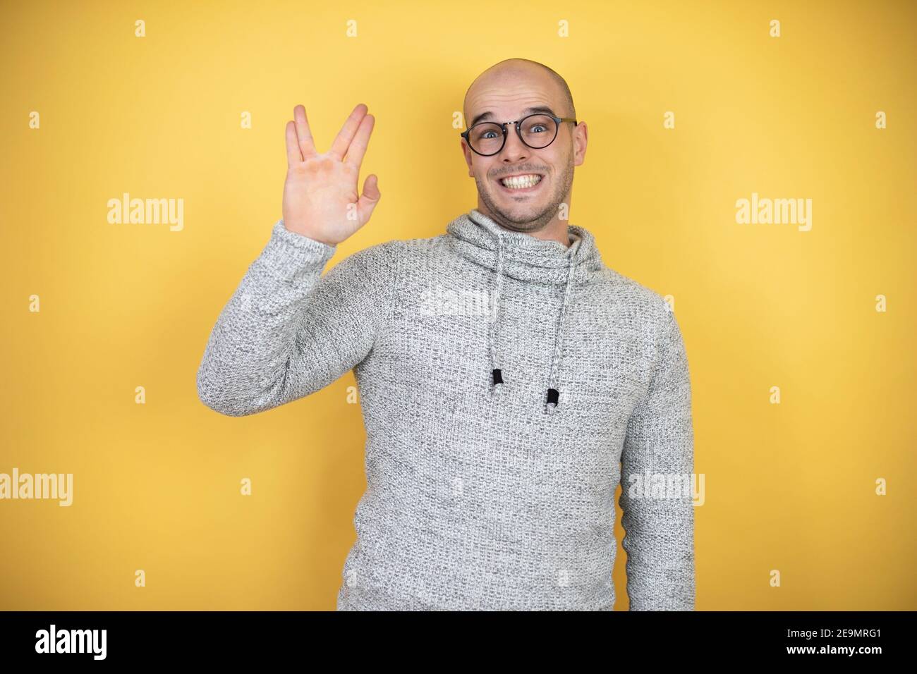 Young bald man wearing glasses over yellow background doing star trek ...