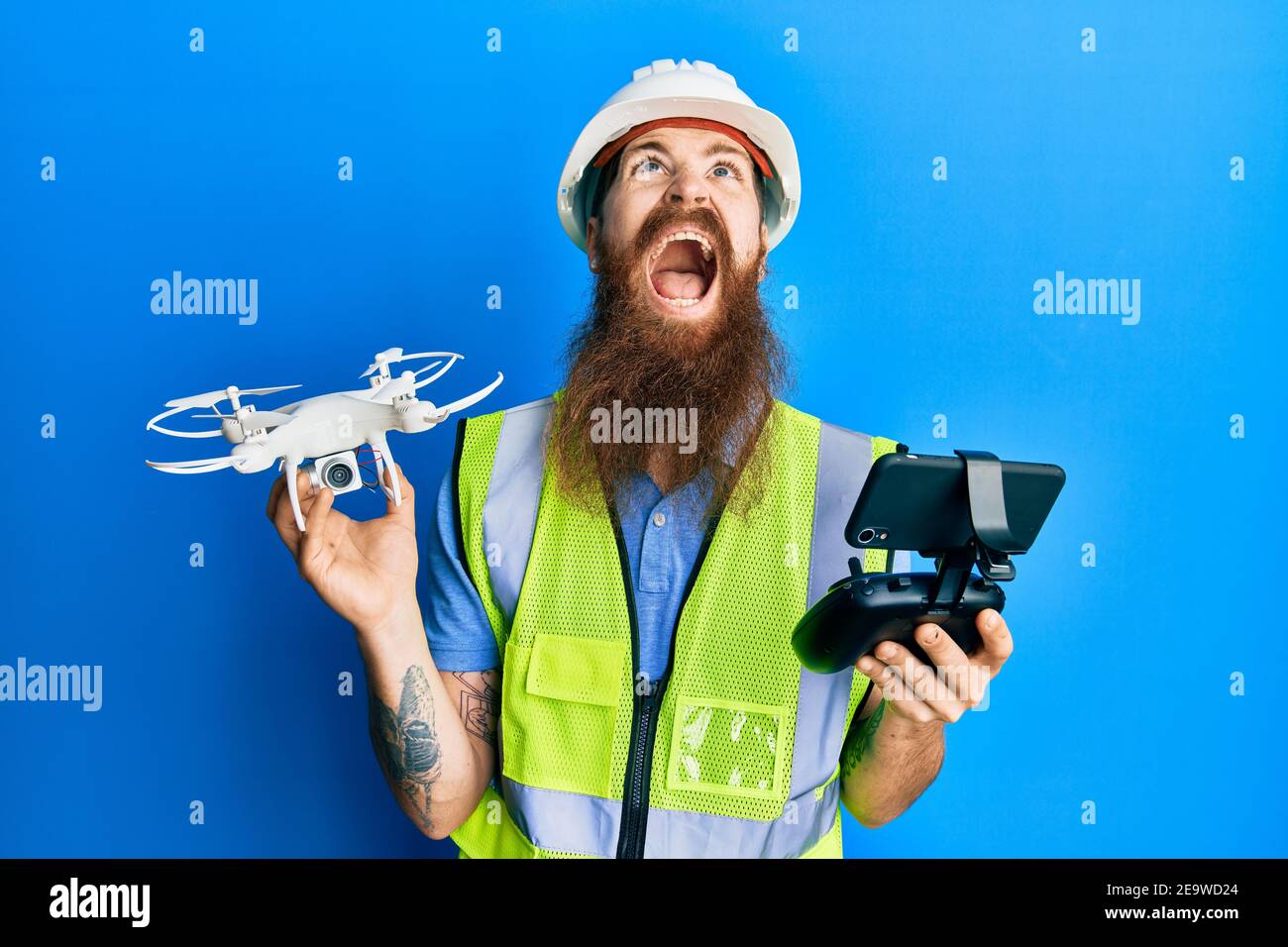 Redhead man with long beard wearing safety hard using drone with remote ...