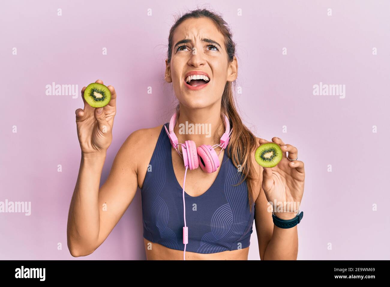 Young beautiful woman wearing gym clothes holding slice of kiwi angry ...