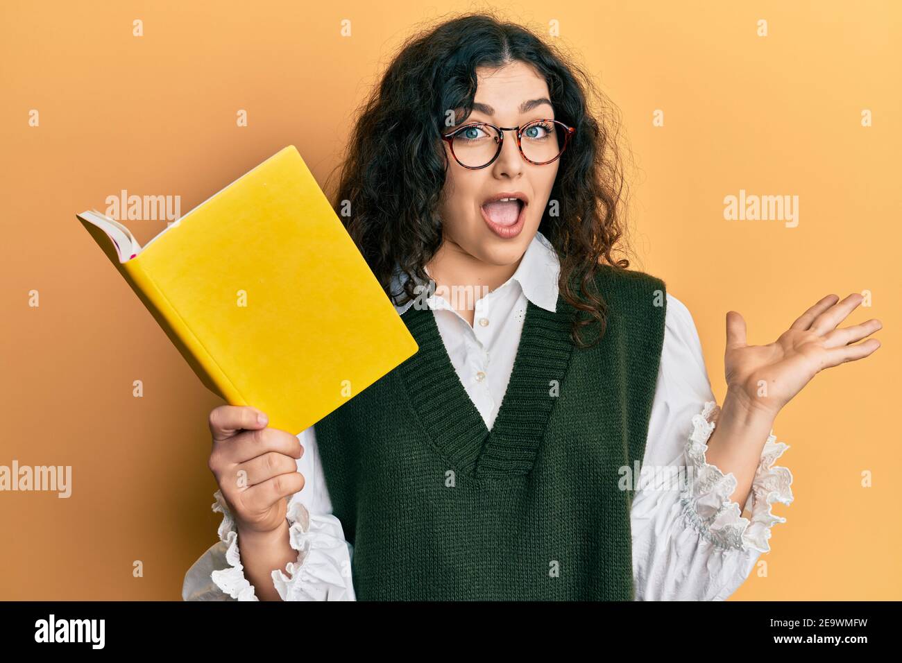 Young brunette woman with curly hair reading a book wearing glasses ...