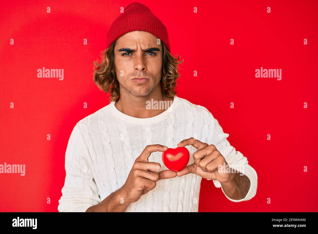 Young hispanic man holding heart skeptic and nervous, frowning upset ...