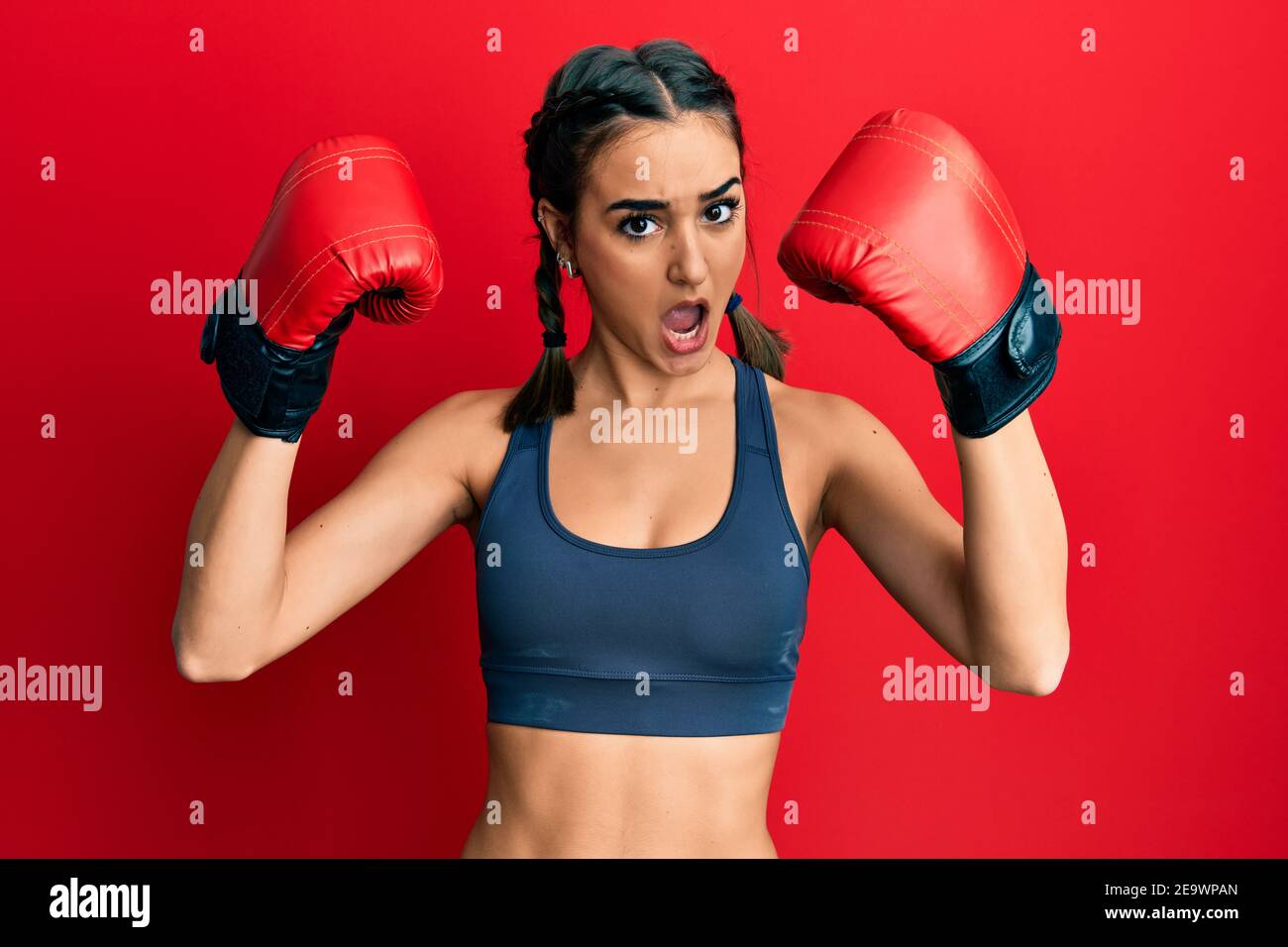 Young brunette girl using boxing gloves wearing braids in shock face ...