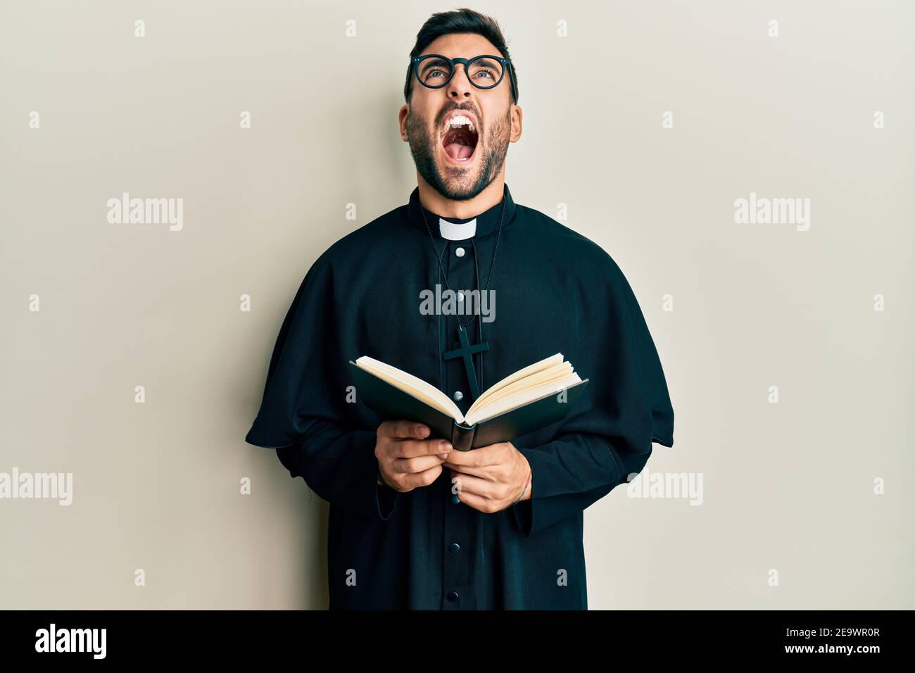 Young hispanic priest man holding bible angry and mad screaming ...