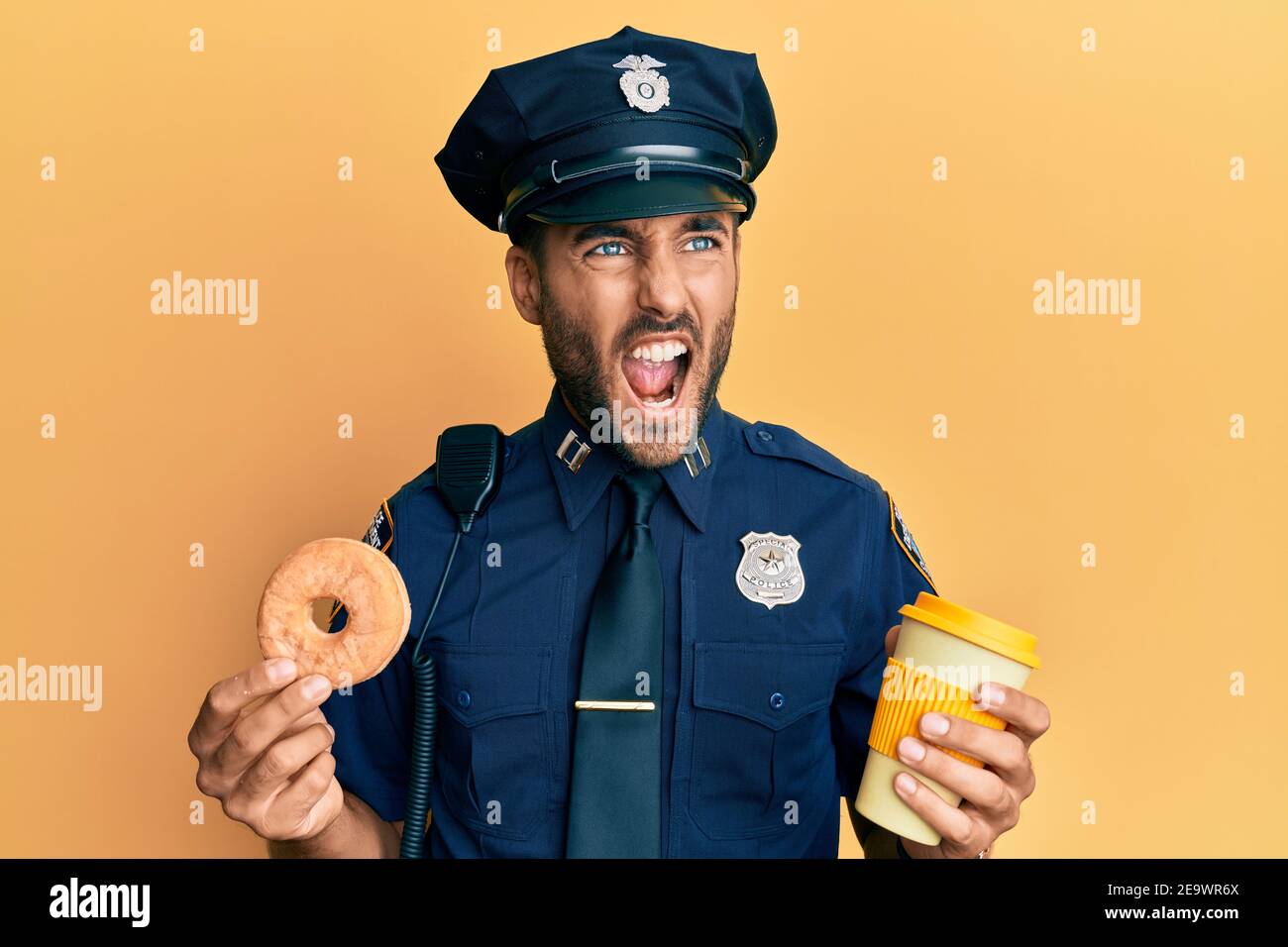 Handsome hispanic police man eating donut and drinking coffee angry and ...