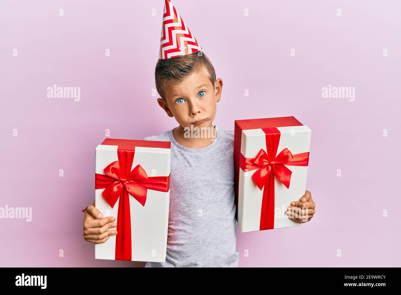 Adorable caucasian kid wearing birthday hat holding presents depressed ...