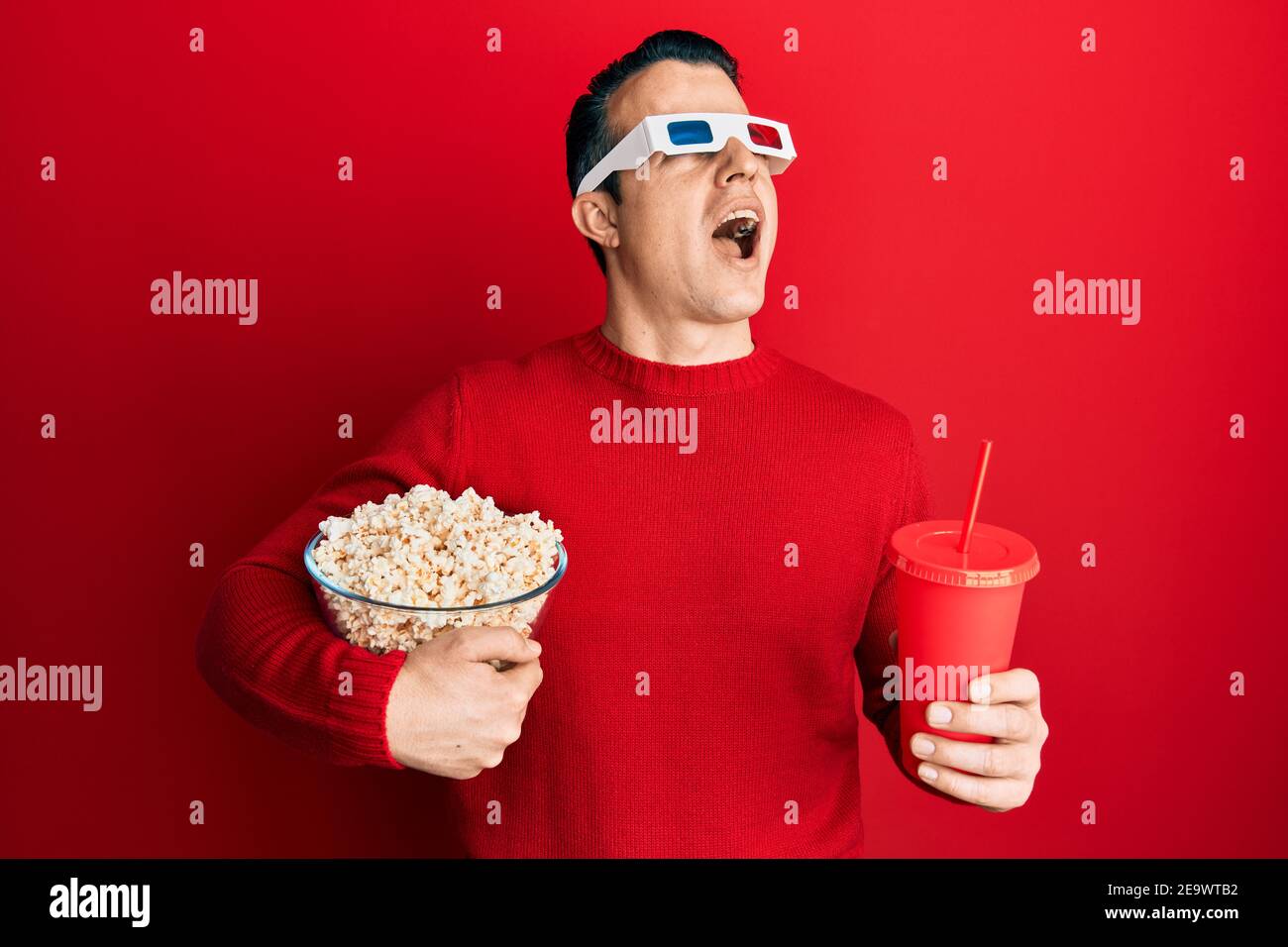 Handsome young man eating popcorn and soda wearing 3d glasses angry and ...