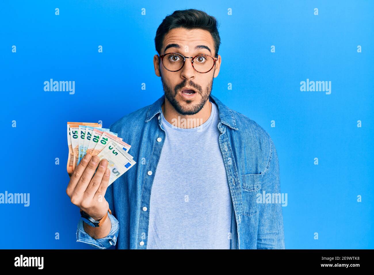Young hispanic man holding euro banknotes scared and amazed with open ...