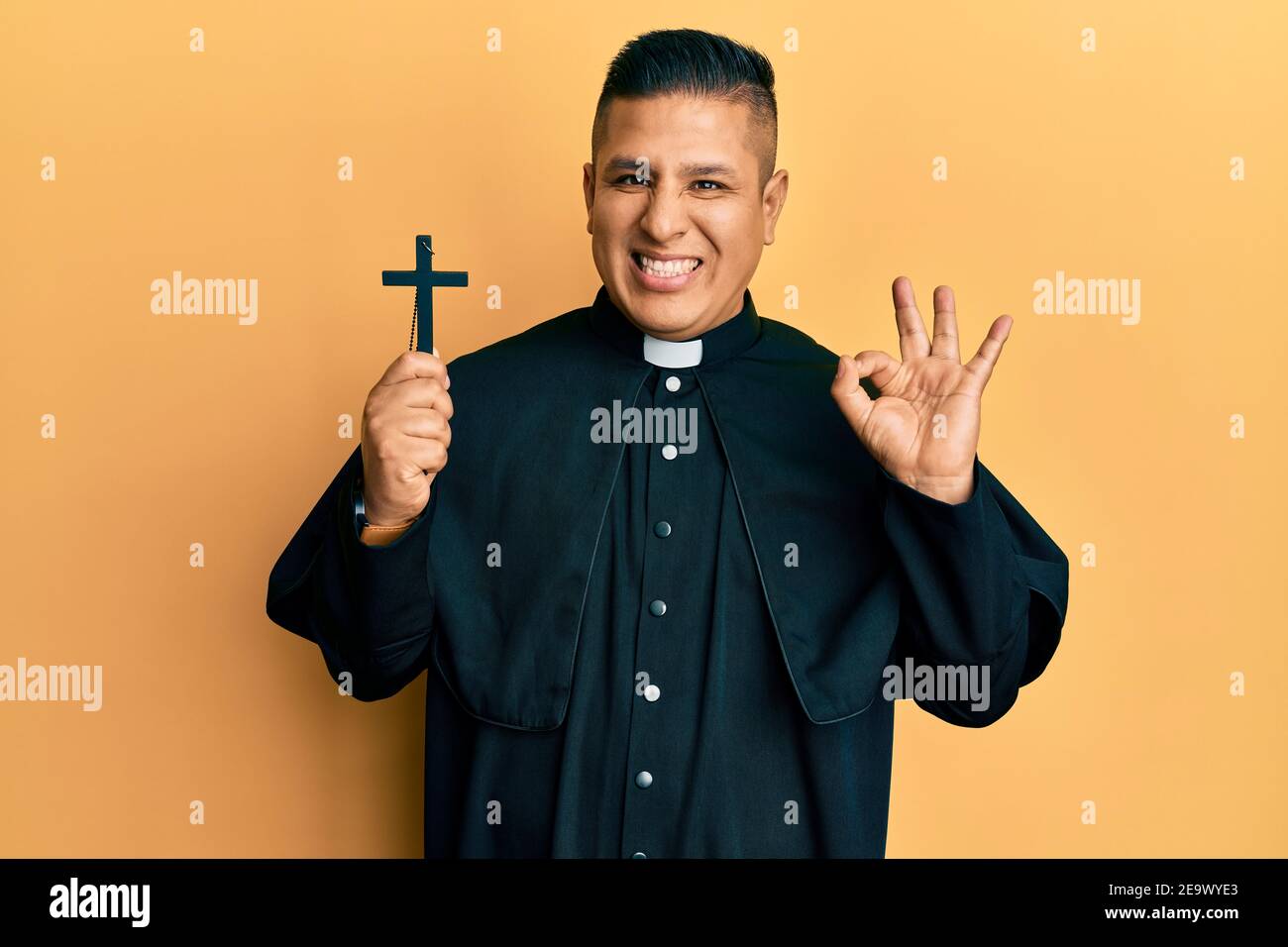 Young latin priest man holding crucifix doing ok sign with fingers ...