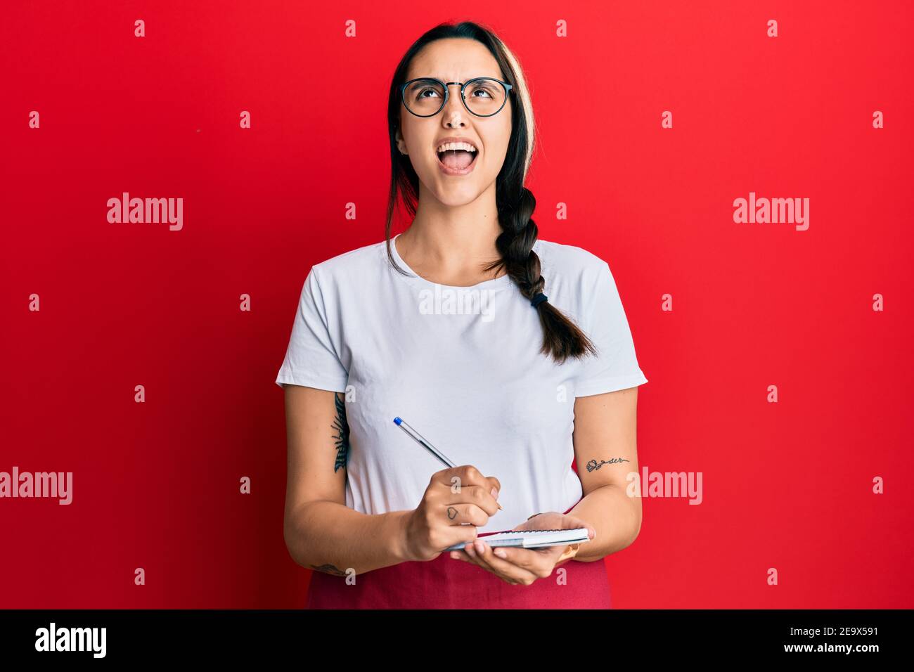 Young hispanic woman wearing waitress apron taking order angry and mad screaming frustrated and ...