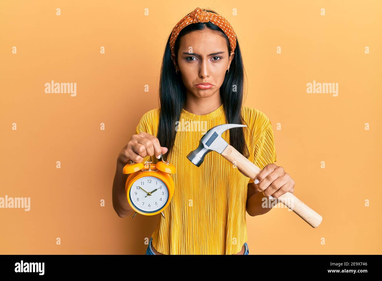 Young brunette woman holding alarm clock and hammer depressed and worry ...