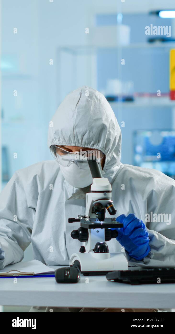 Biologist in ppe suit working on microscope and writing information on ...