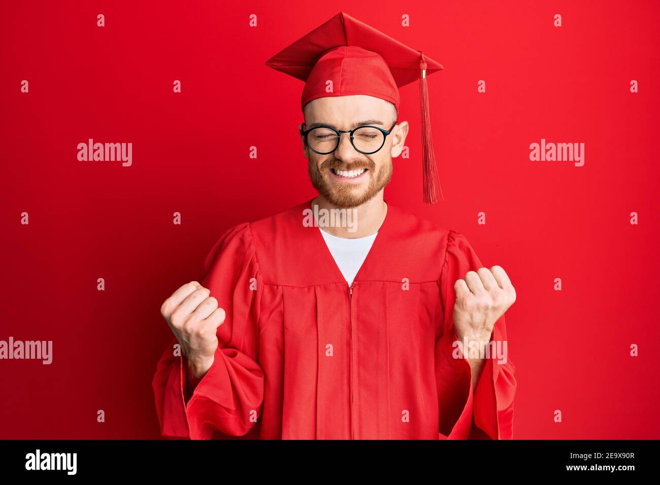 Young redhead man wearing red graduation cap and ceremony robe excited ...