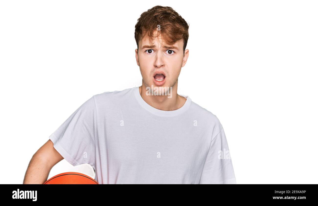 Young caucasian man holding basketball ball scared and amazed with open ...