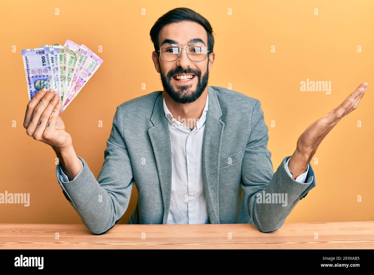 Young hispanic man holding indian rupee banknotes sitting on the table ...