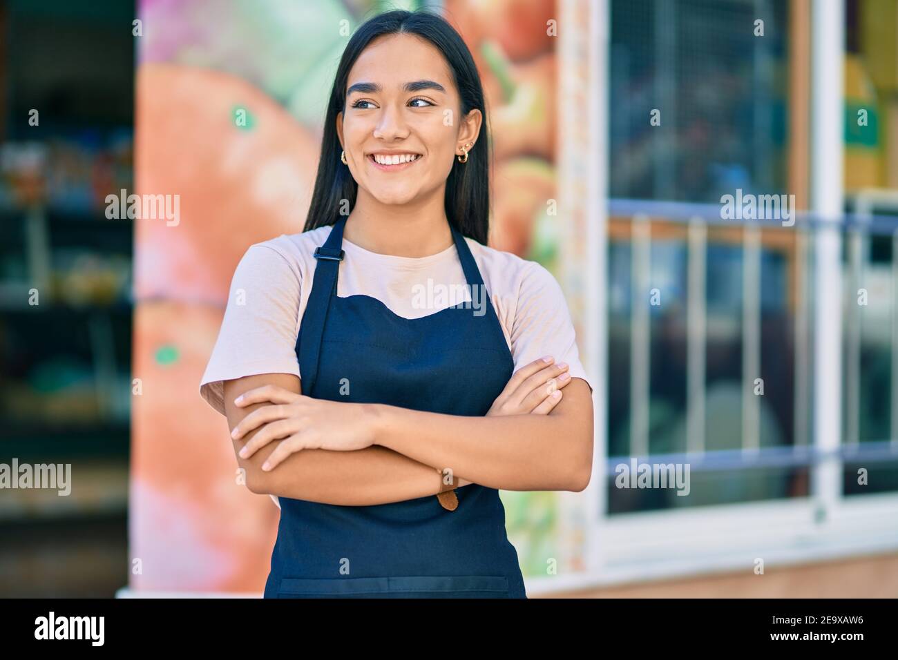 Young latin shopkeeper girl with arms crossed smiling happy at the ...