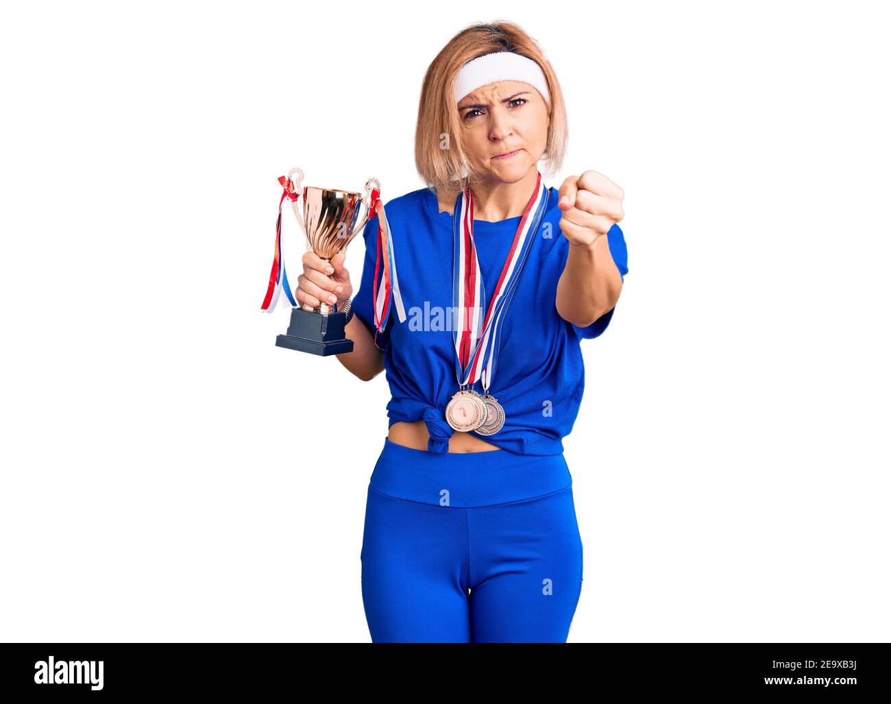 Young blonde woman holding champion trophy wearing medals annoyed and ...