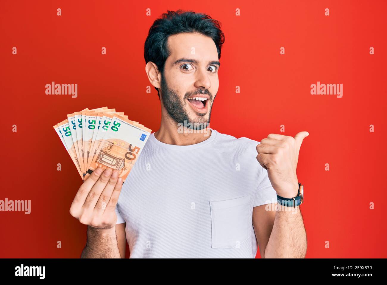 Young hispanic man holding euro banknotes pointing thumb up to the side ...