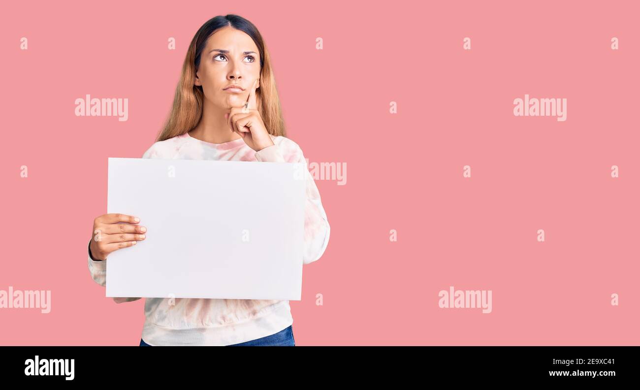 Beautiful young woman holding blank empty banner serious face thinking ...