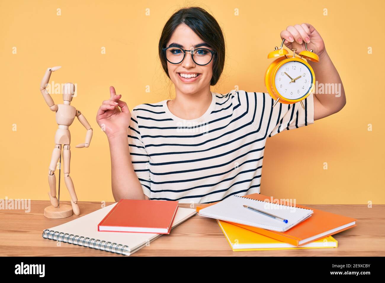 Brunette teenager girl holding alarm clock studying for exam smiling ...