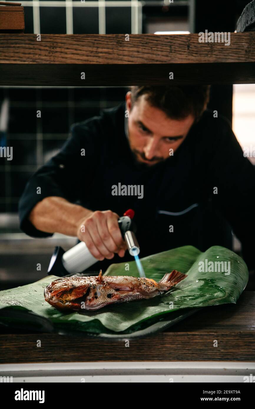 Concentrated male chef cooking fried fish with gas torch while working ...