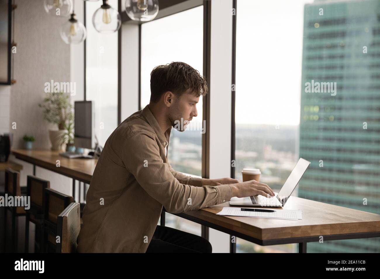 Concentrated millennial man using laptop at desk near panoramic window ...