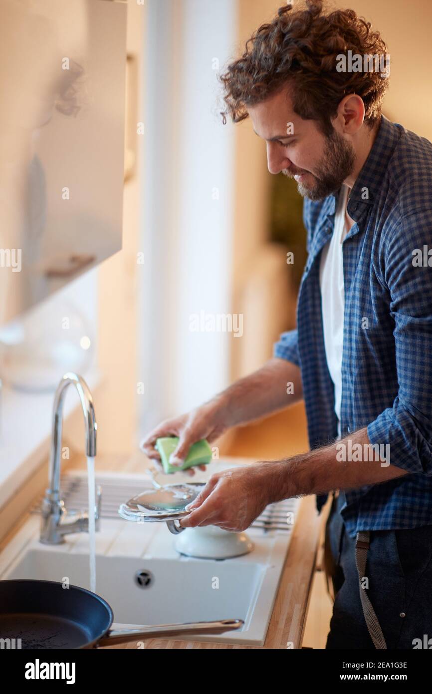 A cheerful young man washing dishes in a relaxed atmosphere in the ...