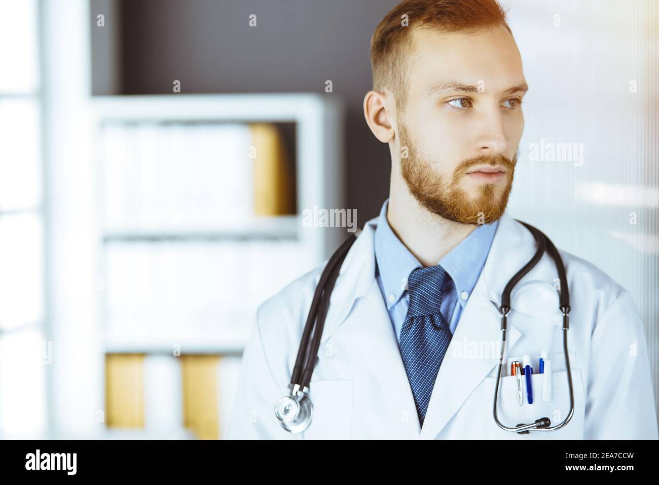 Red-bearded doctor standing straight in sunny clinic near his working ...